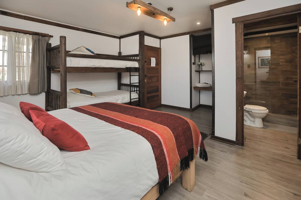 Quarto com beliche, cabeceira de madeira, travesseiro vermelho e cobertor. Há um banheiro com vaso sanitário e espelho.