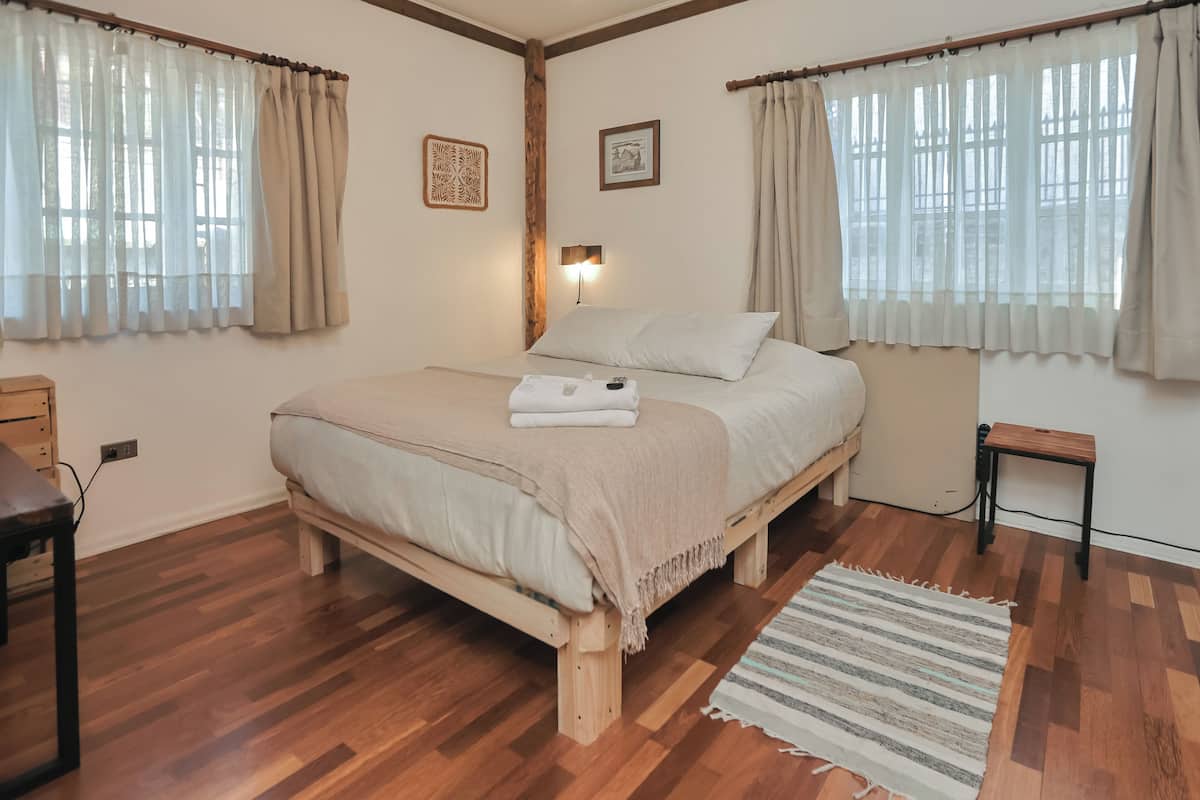 Quarto com cama de madeira, mesa de cabeceira, abajur e janela com cortinas.
