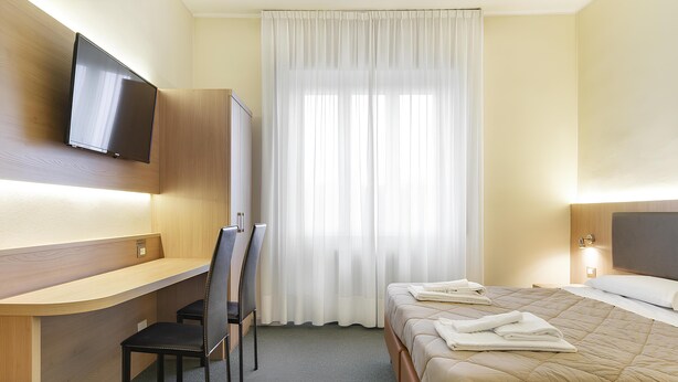 Quarto de hotel com escrivaninha, cadeira, cama e televisão fixada na parede.
