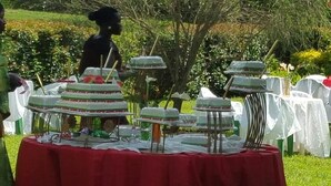 Mesa com bolos dispostos em camadas e uma pessoa ao fundo.