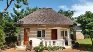 Uma casa com telhado de palha, varanda branca, portas de madeira e um pequeno alpendre.