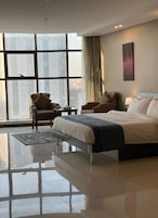 Quarto de hotel moderno com uma cama grande, duas poltronas, uma mesa pequena e vista para a cidade através de grandes janelas.