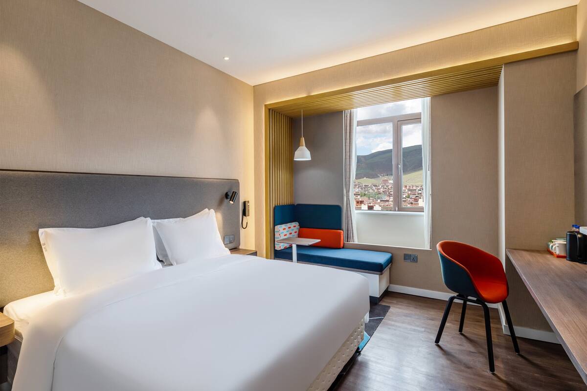 Quarto de hotel com uma cama grande, uma escrivaninha com cadeira e uma janela com vista para a cidade.