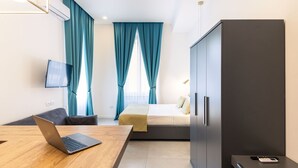 Quarto de hotel moderno com escrivaninha de madeira, um laptop, cama com manta amarela, cortinas verde-água e um guarda-roupa grande.