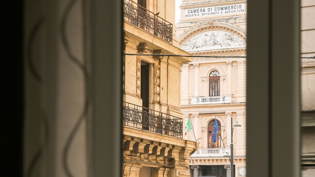 Vista pela janela de um edifício com varanda, rua com uma scooter estacionada e uma bandeira.