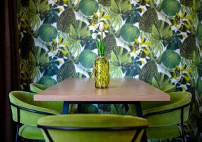 Uma mesa com um vaso de flores, cadeiras verdes e uma parede com padrão de folhas tropicais.