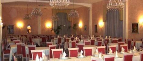 Salão de banquetes com várias mesas preparadas para refeições, cada uma com toalhas brancas e cadeiras vermelhas.
