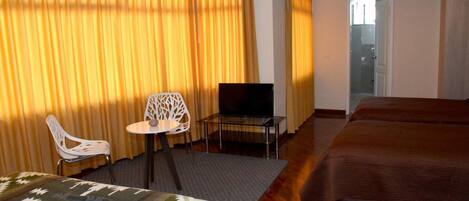 Quarto de hotel com cama, televisão, pequena mesa com cadeira e janela com cortinas.