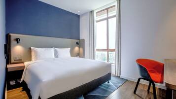 Quarto de hotel com cama grande, mesa de cabeceira, uma cadeira e janela com cortinas.