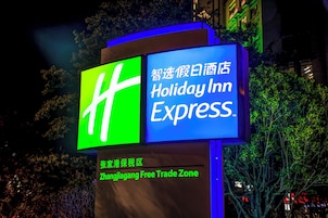 Placa do Holiday Inn Express com esquema de cores verde e azul, localizada em área comercial com árvores e edifícios ao fundo.