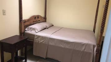 Quarto pequeno com uma cama de solteiro, cabeceira e quatro suportes de madeira. Há uma pequena mesa de madeira ao lado da cama.