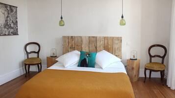 Quarto com cabeceira de madeira, uma cama grande com manta amarela mostarda, duas mesinhas de cabeceira de madeira com luminárias, e duas poltronas estofadas.