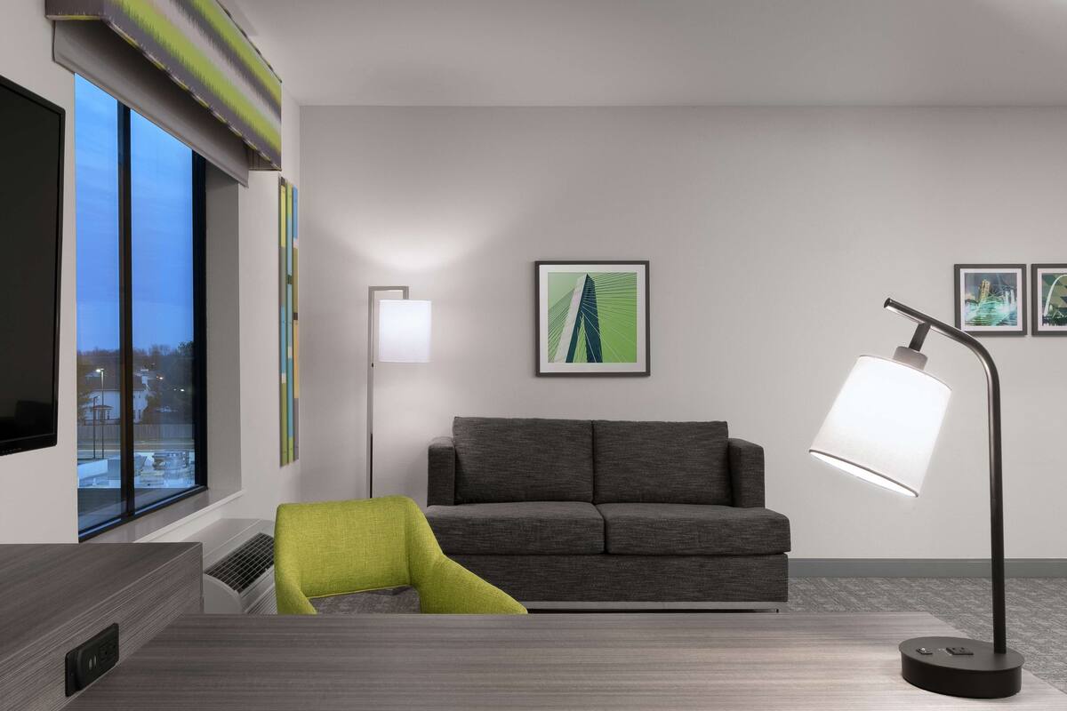 Quarto de hotel moderno com um sofá cinza, uma poltrona verde, um abajur de chão e uma grande janela com vista para a cidade.