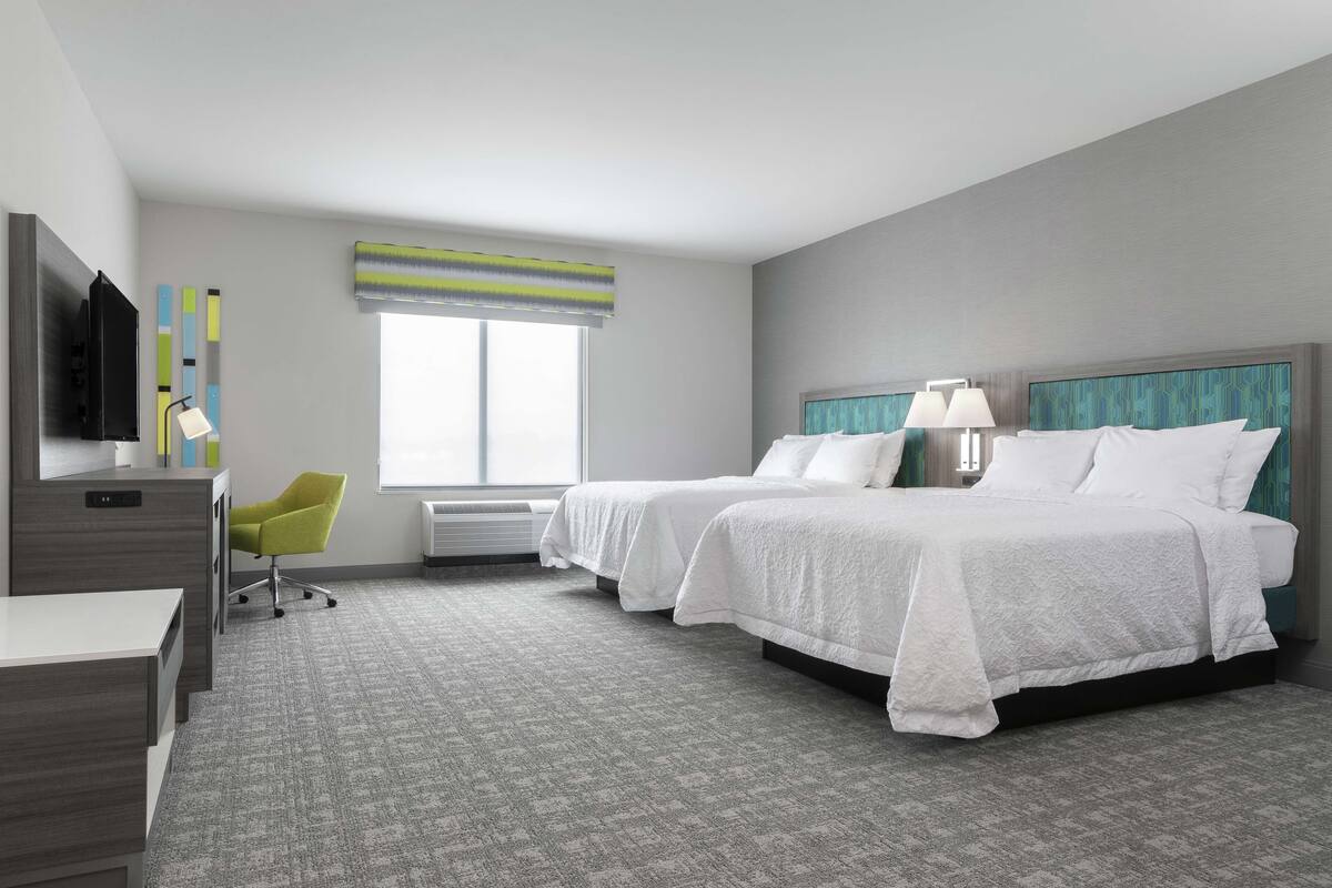 Quarto de hotel com duas camas, uma escrivaninha, uma cadeira, uma televisão e uma janela com vista.
