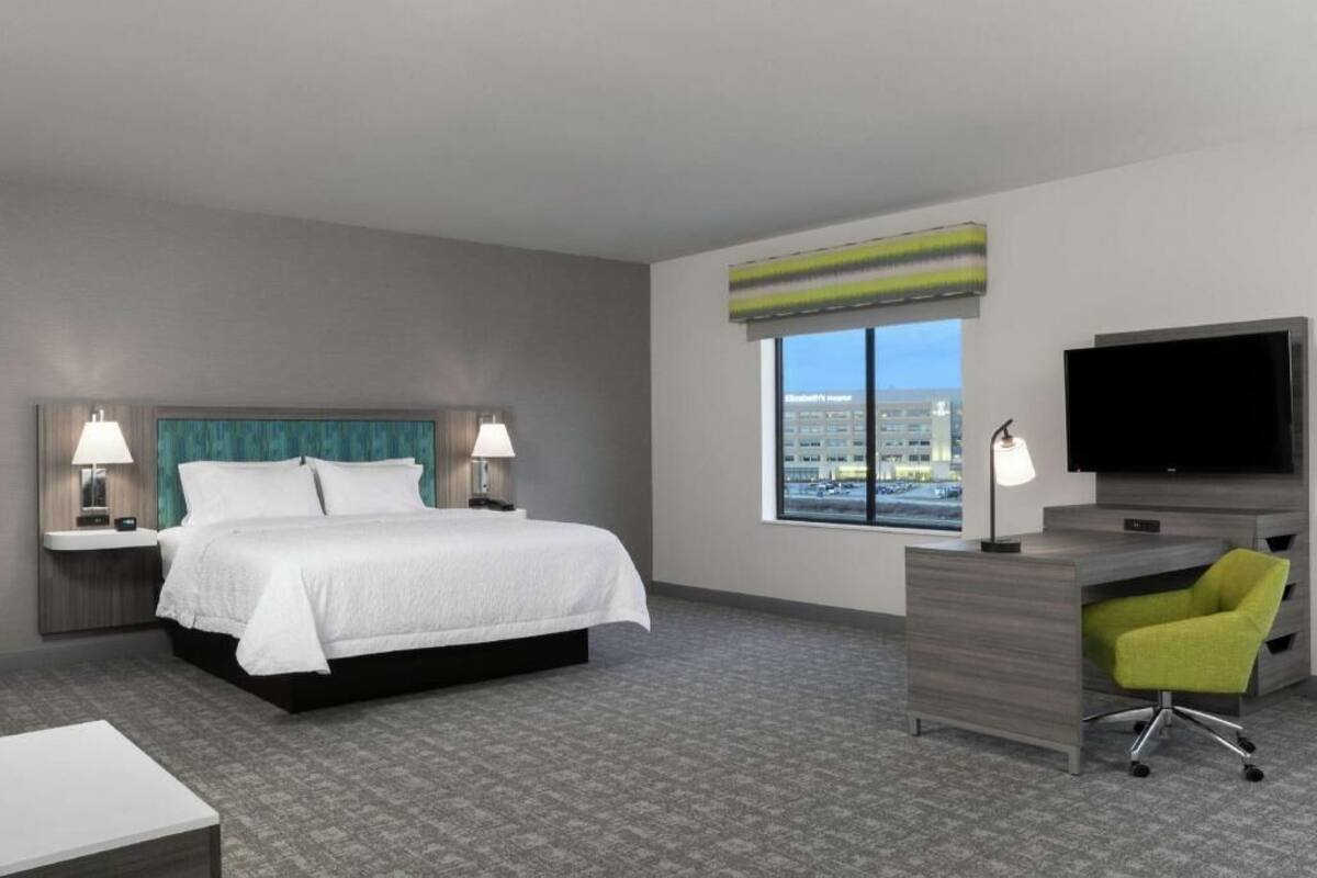 Quarto de hotel com cama, escrivaninha, cadeira, televisão, janela e abajur.