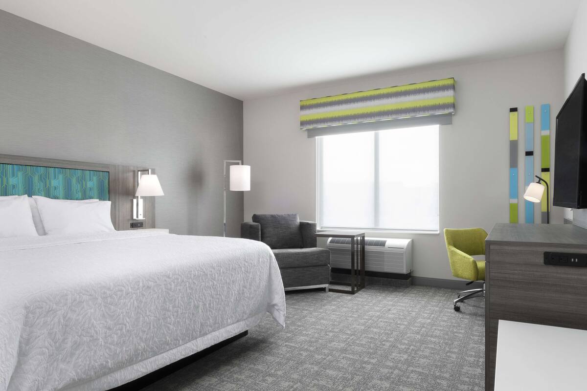 Quarto de hotel com cama grande, escrivaninha, cadeira, janela com vista e televisão presa à parede.