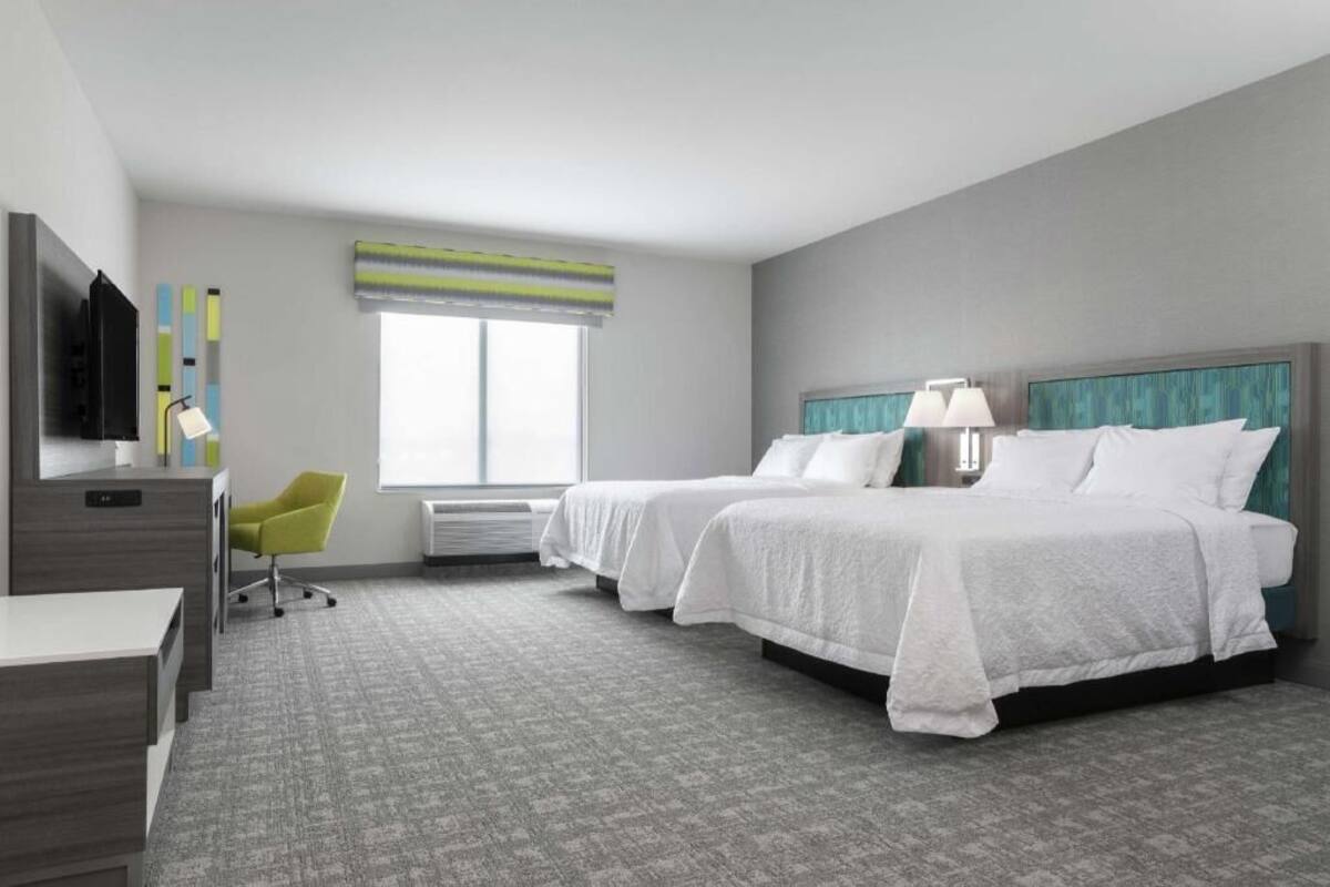 Quarto de hotel com duas camas, uma escrivaninha, uma cadeira, uma televisão e janela com persianas.