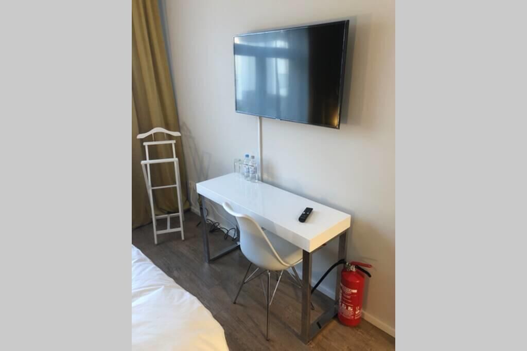Quarto de hotel com escrivaninha branca, televisão fixada na parede, cadeira branca, escada e extintor de incêndio vermelho.