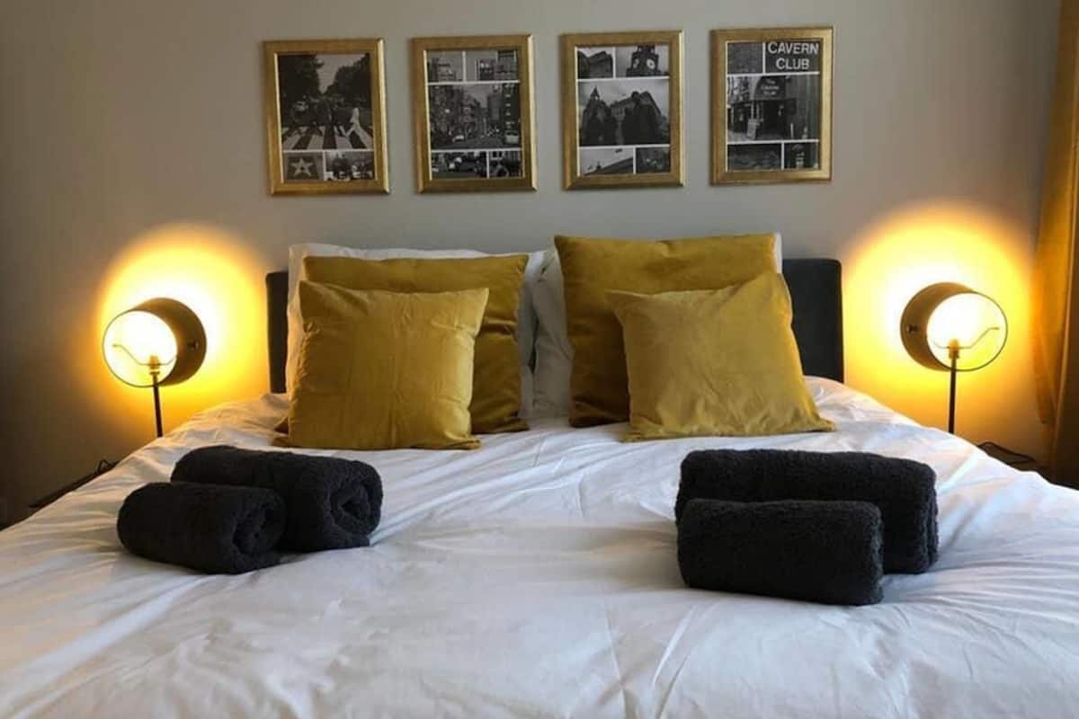 Cama bem arrumada com lençóis brancos e duas almofadas, um par de toalhas enroladas e duas luminárias na parede.