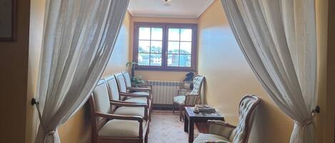 Um quarto com piso de madeira, uma janela com cortinas e várias cadeiras dispostas ao redor de uma mesa.