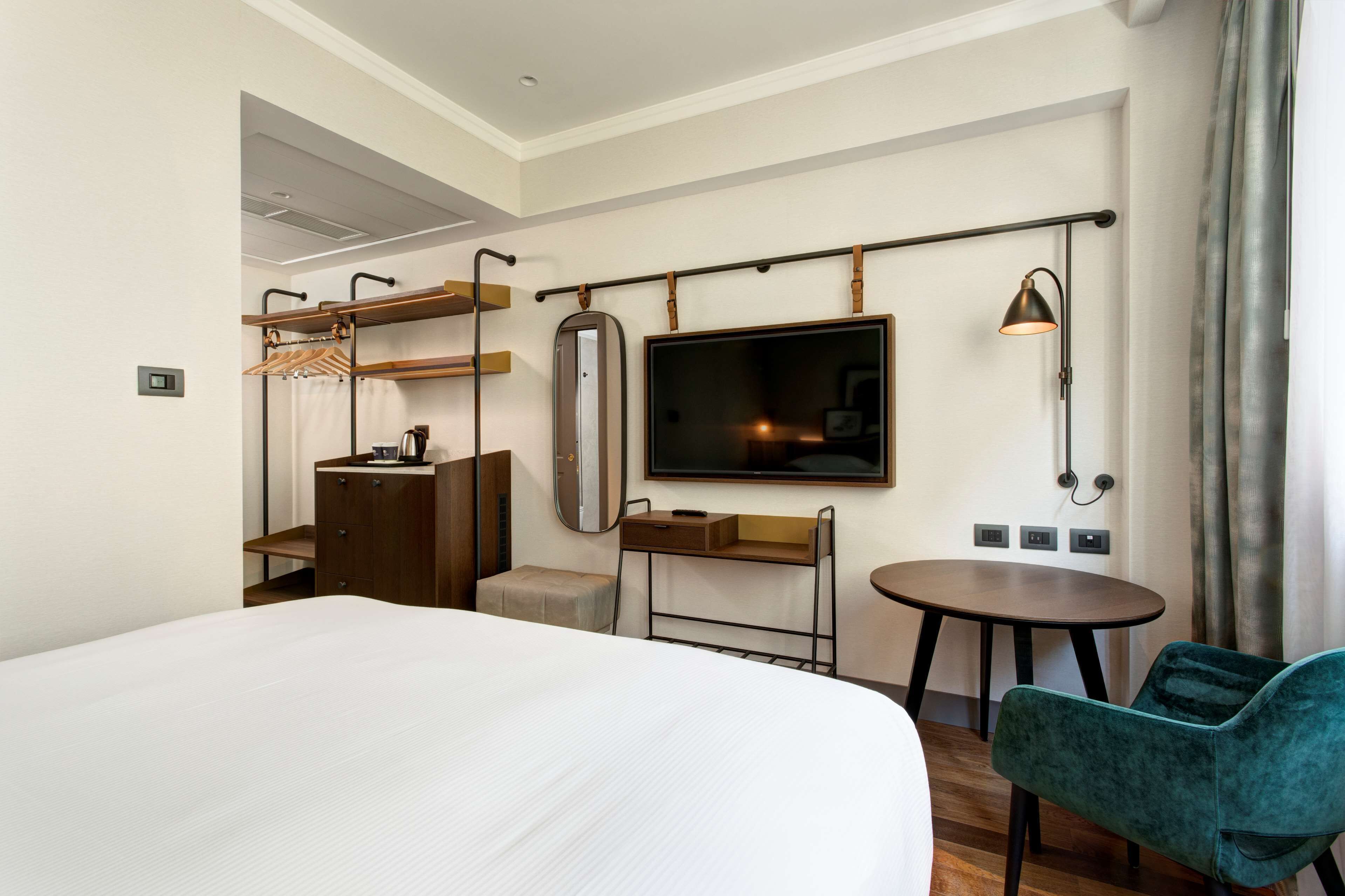 Quarto de hotel com uma cama grande, uma escrivaninha com cadeira, televisão na parede, uma mesinha e um guarda-roupa.