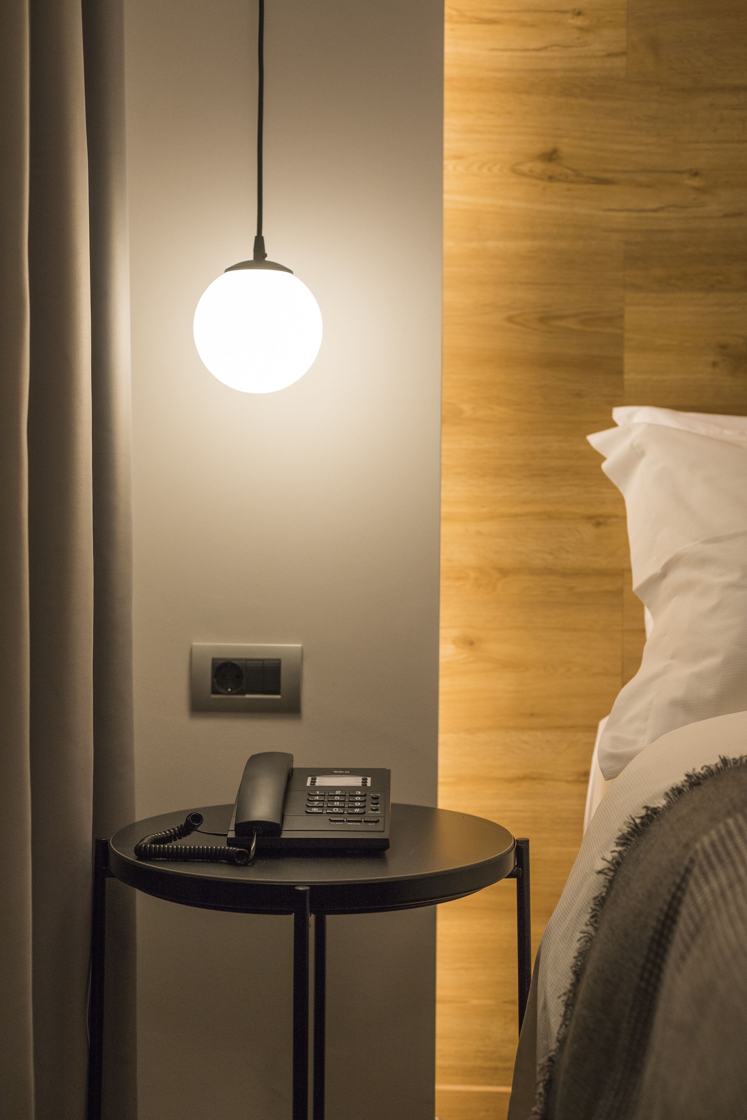 Quarto de hotel com uma cama, uma mesa de cabeceira com telefone e um candeeiro suspenso.