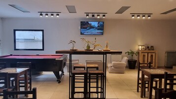 Uma mesa de bilhar, banquetas de bar, uma televisão na parede e um vaso de planta.