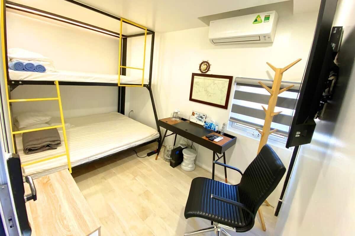 Quarto compacto com beliches, uma escrivaninha, uma cadeira, ar condicionado e janela com persianas.