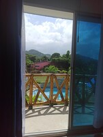 Vista através de uma porta de vidro para uma área de piscina com cerca de bambu, um edifício vermelho e uma paisagem montanhosa.