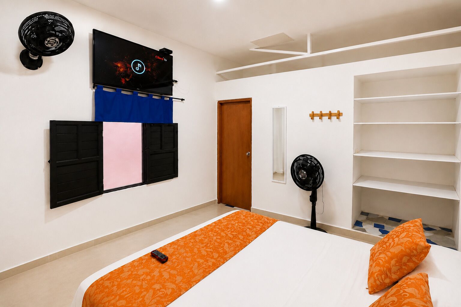 Um quarto com cama, TV fixada na parede, guarda-roupa e dois ventiladores.