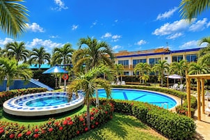 Área de piscina de hotel com piscina circular, palmeiras e um edifício com fachada azul e branca.