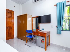 Quarto compacto com guarda-roupa de madeira, escrivaninha com cadeira, pequeno frigorífico, televisão de ecrã plano e janela com cortinas.
