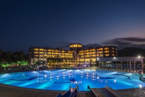 Hotel com piscina com desenhos de golfinhos, cercado por área externa bem iluminada com espreguiçadeiras e um cenário montanhoso.