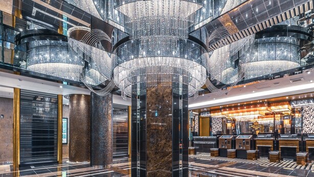 Um lobby de hotel luxuoso com um lustre imponente, colunas de mármore e uma área de recepção moderna.