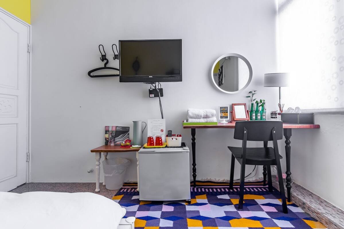 Quarto de hotel com cama, escrivaninha com computador, televisão, espelho e pequeno frigorífico.