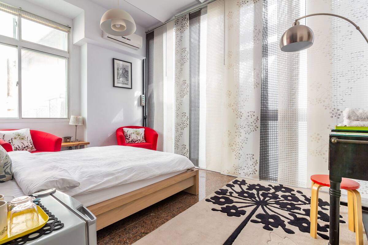 Quarto de hotel moderno com uma cama grande, cadeiras vermelhas, uma mesa com abajur e vista para a cidade através de amplas janelas.