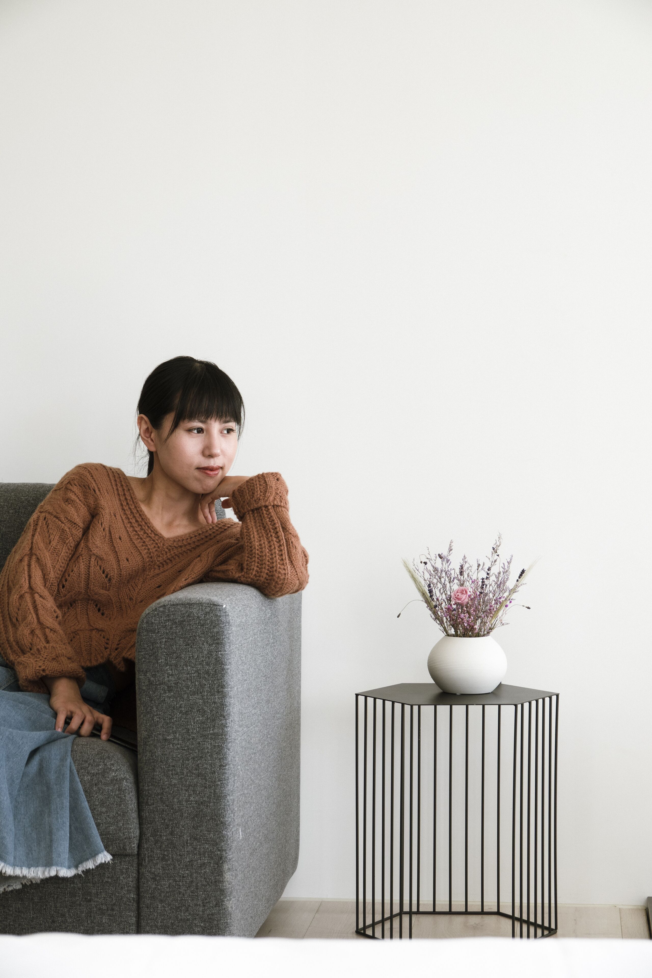 Uma pessoa sentada em um sofá cinza, com um vaso branco de flores sobre uma mesa lateral preta.