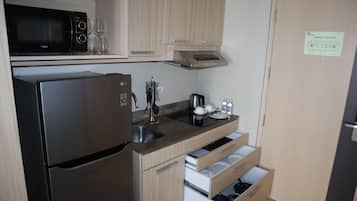 Uma cozinha compacta com frigorífico em aço inoxidável, micro-ondas e lava-loiça. Existem armários com gavetas e uma bancada com chaleira e chávenas.
