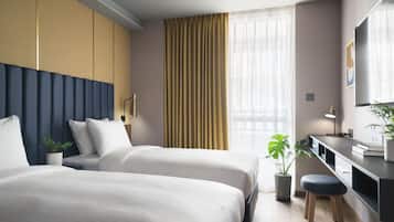 Quarto de hotel com duas camas, uma escrivaninha, uma cadeira, um vaso de plantas e uma grande janela com cortinas.