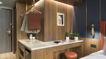 Quarto de hotel moderno com parede de madeira, uma secretária com máquina de café, uma toalha pendurada e uma cama com almofada vermelha.