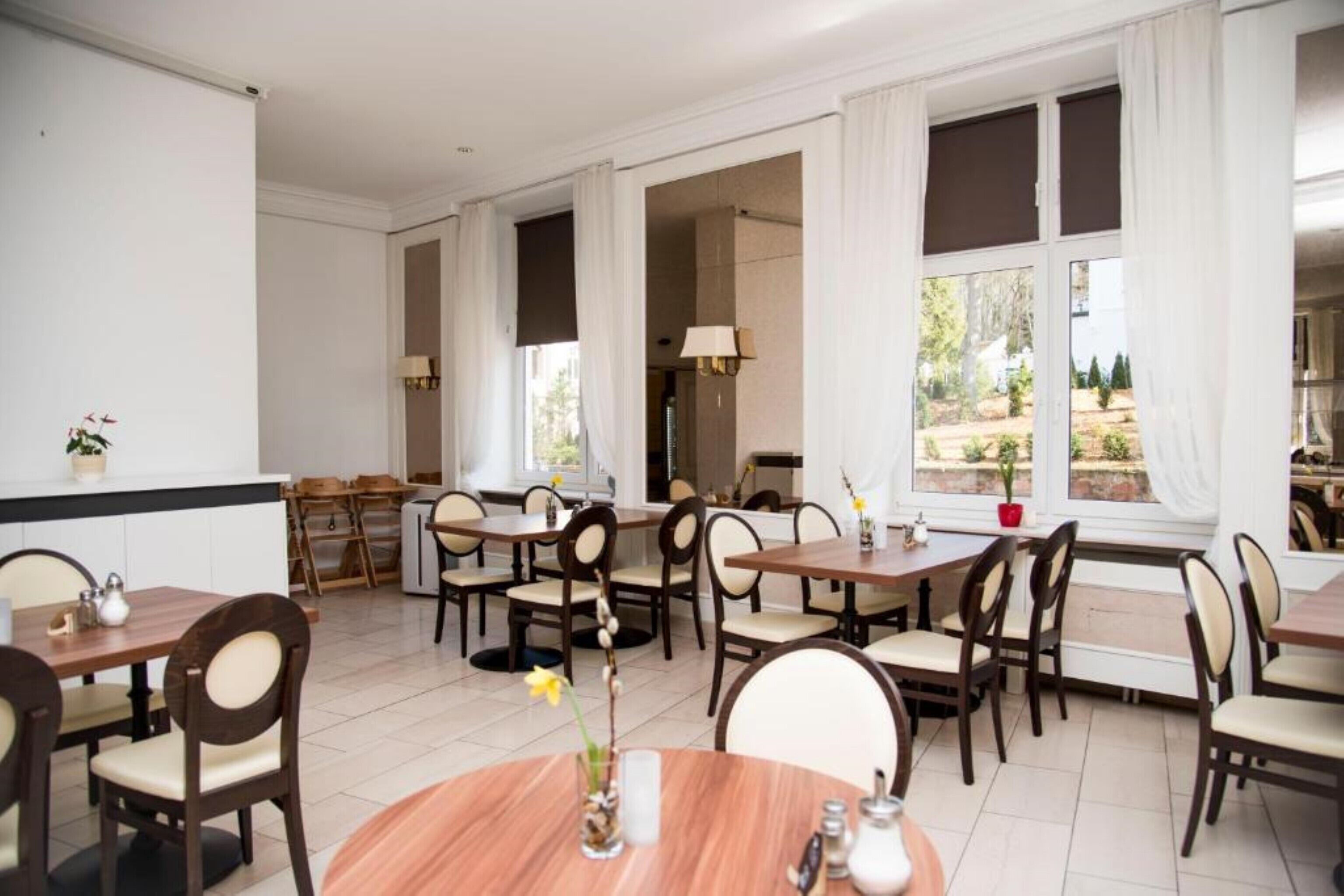 Uma área de refeições com várias mesas e cadeiras, grandes janelas com cortinas e vista para o exterior.