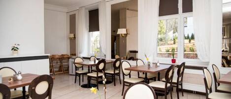 Uma área de refeições com várias mesas e cadeiras, grandes janelas com cortinas e vista para o exterior.