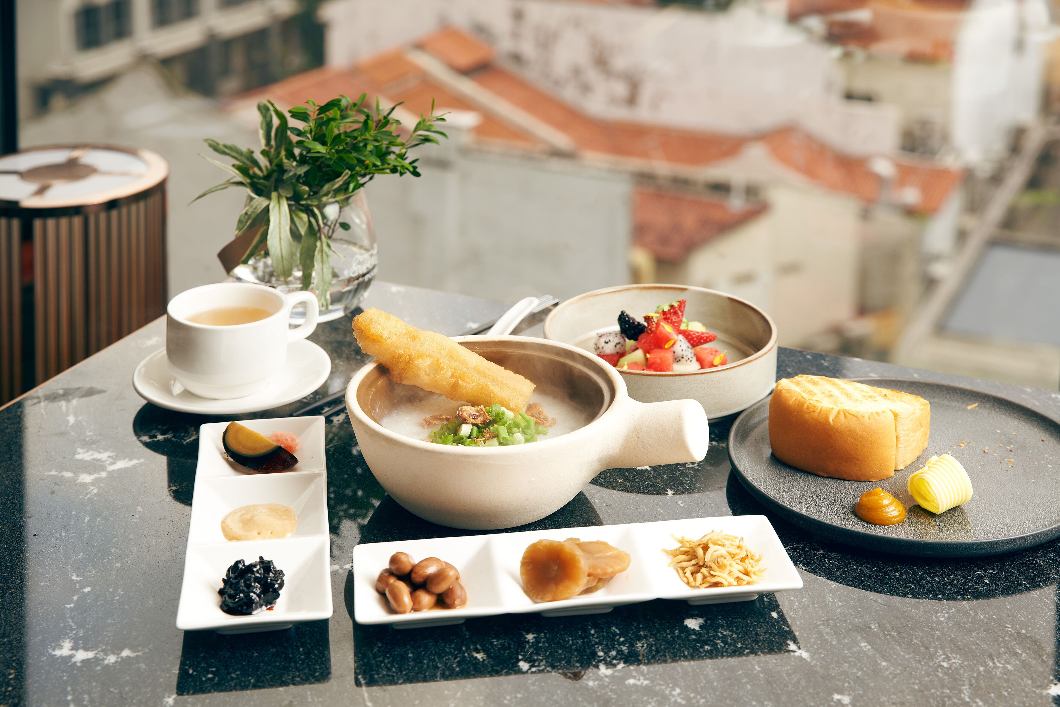 Uma mesa com diversos pratos, incluindo sopa, pão e uma chávena de chá, numa varanda com vista para edifícios.
