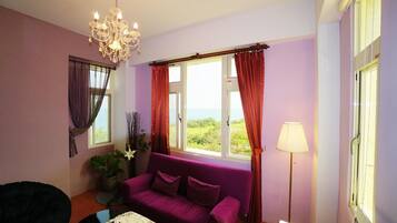 Quarto com sofá roxo, um candelabro, uma mesa com um vaso de flores e vista para o exterior através de grandes janelas.