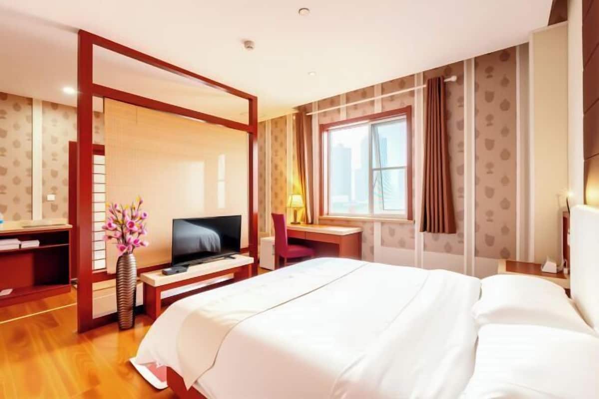 Quarto de hotel com cama, escrivaninha com televisão, cadeira e janela com cortinas.