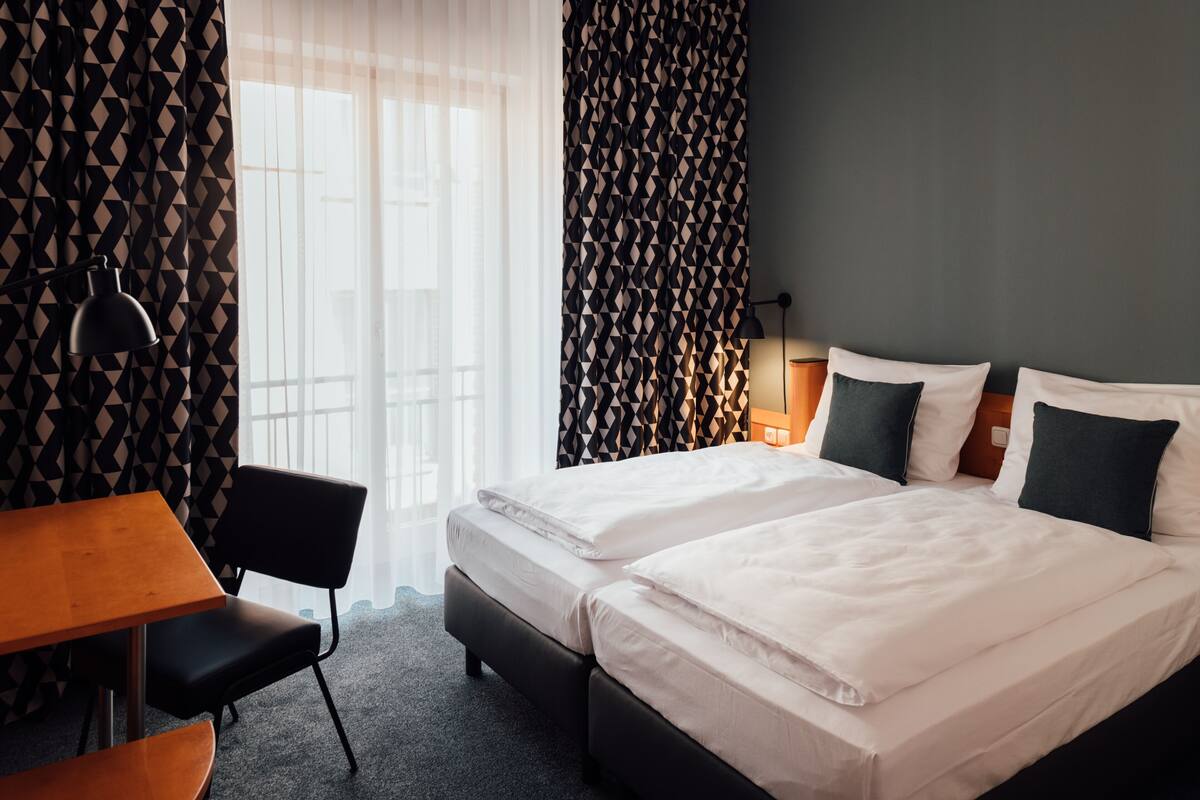 Quarto de hotel com duas camas, uma mesa, uma cadeira e janela com cortinas.