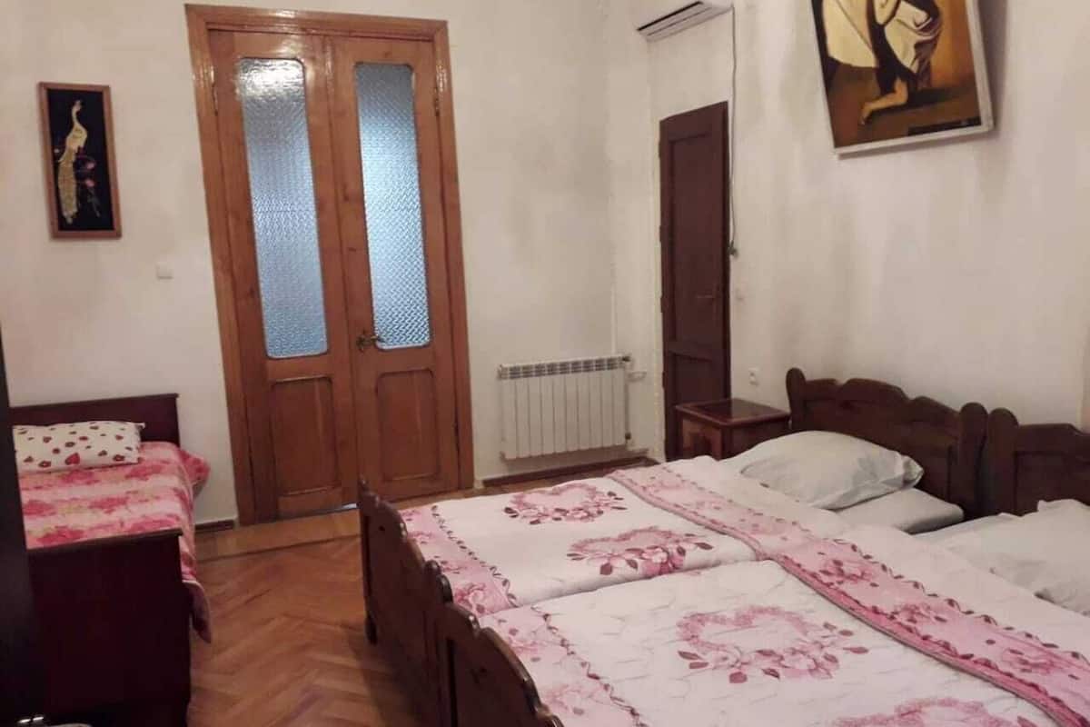Quarto com cama de madeira, mesa de cabeceira de madeira, cômoda, porta com painel de vidro fosco e radiador.