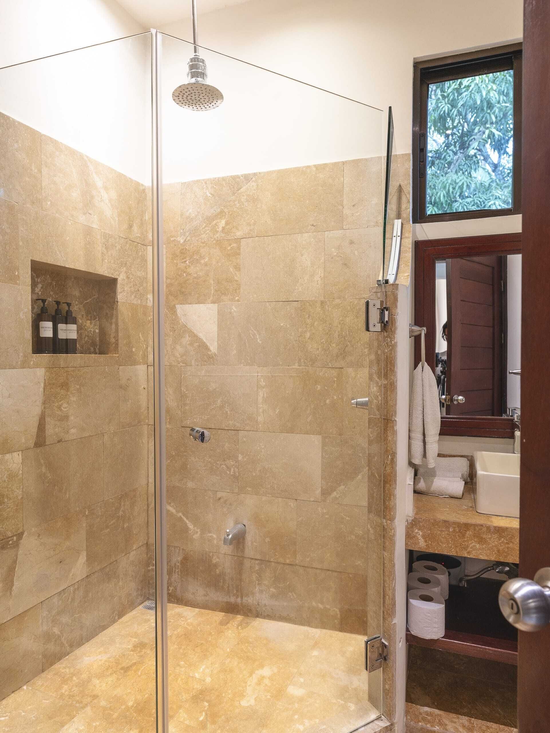 Um chuveiro com paredes de vidro, azulejos bege, um nicho pequeno com artigos de higiene e vista para o banheiro por uma janela.