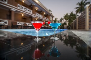 Dois cocktails sobre uma superfície refletora perto de uma área de piscina, com um edifício ao fundo.