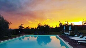 Piscina com espreguiçadeiras e guarda-sóis, cercada por árvores, ao pôr do sol.
